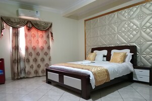 Quarto de hotel com uma cama grande, cabeceira com padrão geométrico, mesa de cabeceira e um refrigerador vermelho.