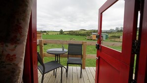 Um deck de madeira com uma mesa e duas cadeiras, com vista para um campo verde e edifícios agrícolas.
