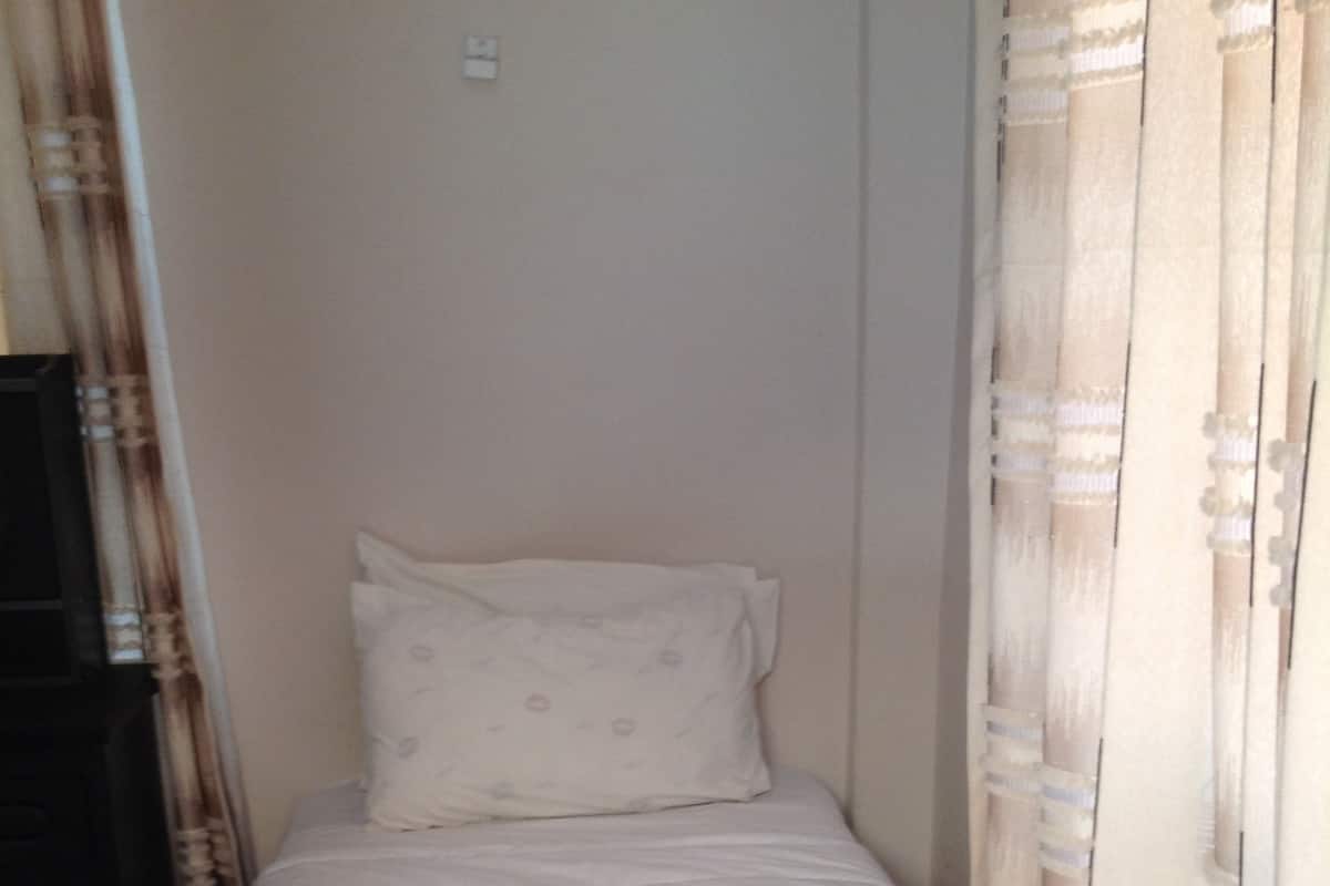 Cama de casal com colchão e almofadas brancas, toalha dobrada e parede lisa com interruptor de luz.
