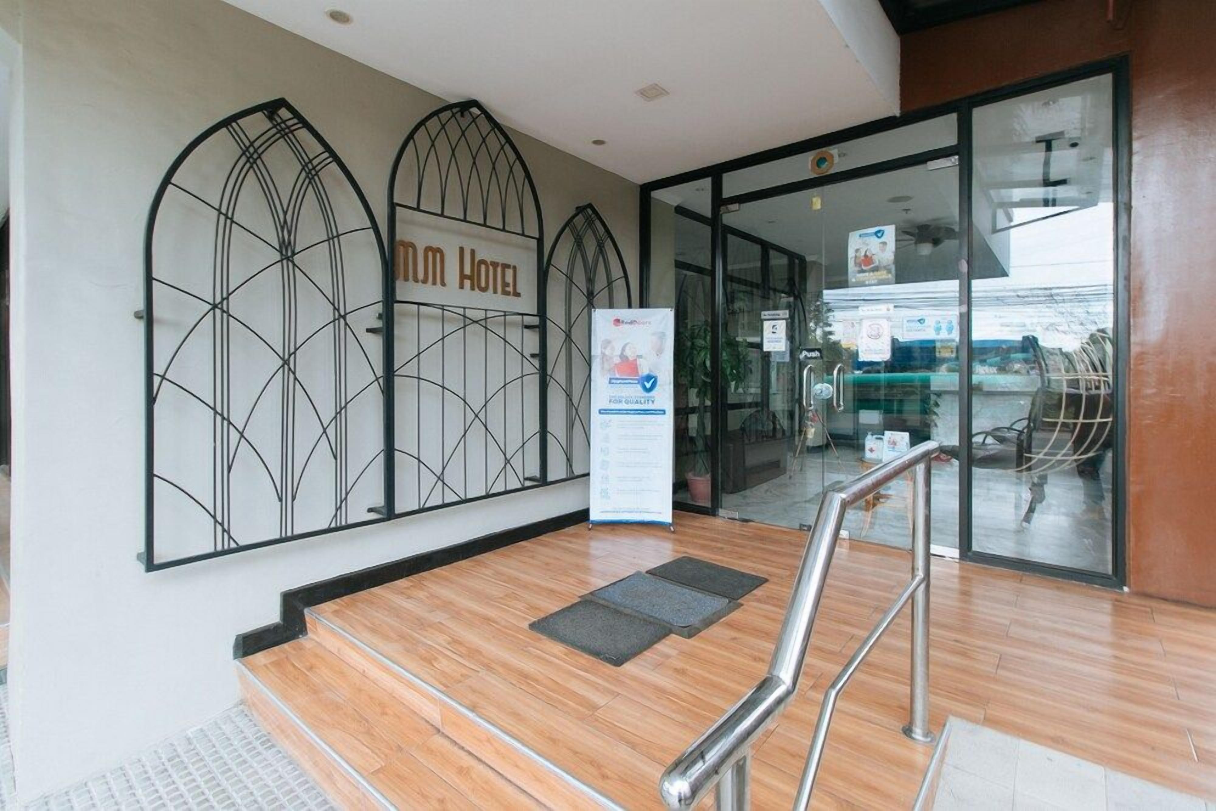 Entrada de um hotel com design moderno, exibindo uma placa, porta de vidro e escadaria de metal que leva à entrada.