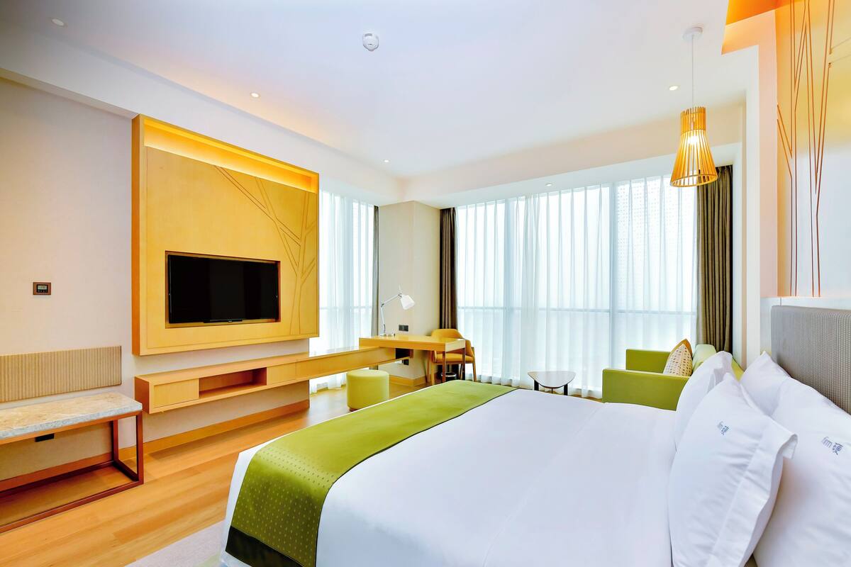 Quarto de hotel moderno com uma cama grande, televisão de tela plana na parede, escrivaninha com cadeira e uma janela ampla com cortinas.