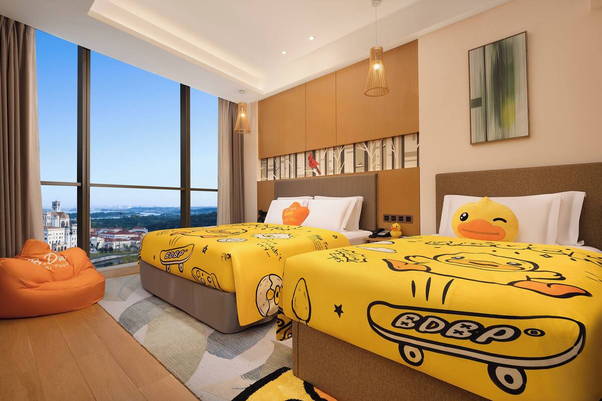 Quarto de hotel com duas camas, uma janela grande, vista para a cidade e um puff.