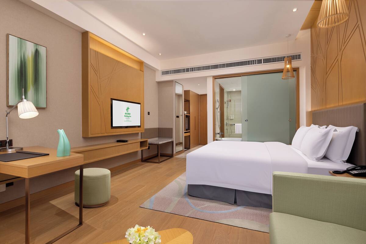 Quarto de hotel moderno com uma cama grande, uma mesa com um computador, uma televisão e um banheiro visível através de uma porta aberta.