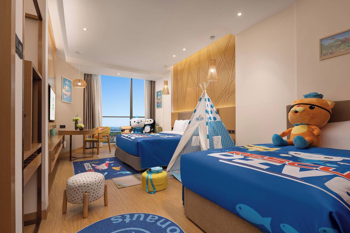 Quarto de hotel com duas camas, uma escrivaninha e uma janela com vista.