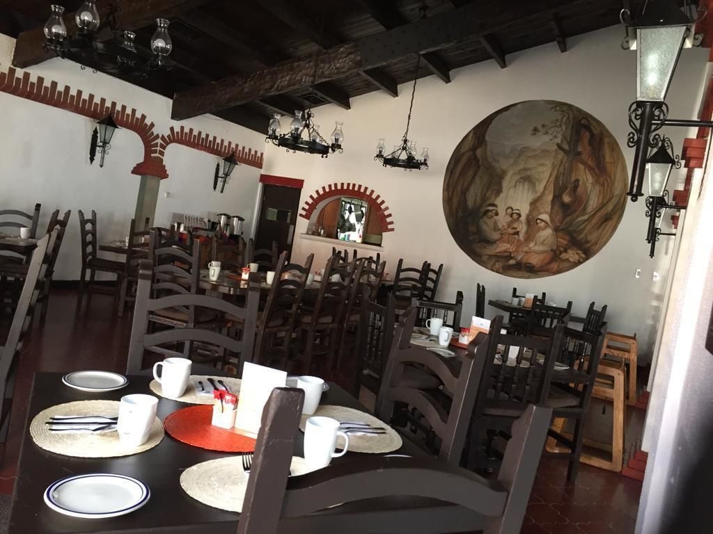Uma sala de jantar com várias mesas e cadeiras, um mural na parede e luminárias pendentes.