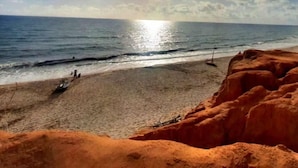 Uma praia com formações rochosas em primeiro plano e céu claro com o sol a brilhar.
