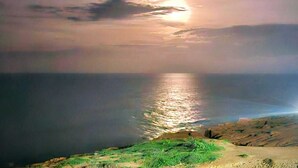 Paisagem costeira com pôr do sol sobre o oceano, litoral rochoso e vegetação escassa.