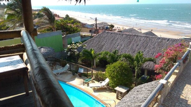 Área de piscina com vista para a praia, cadeiras de descanso e uma construção com telhado de palha.