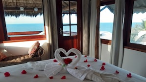 Uma cama com toalha disposta em formato de coração e pétalas de rosa, uma cadeira de vime e vista para o oceano através de uma janela.