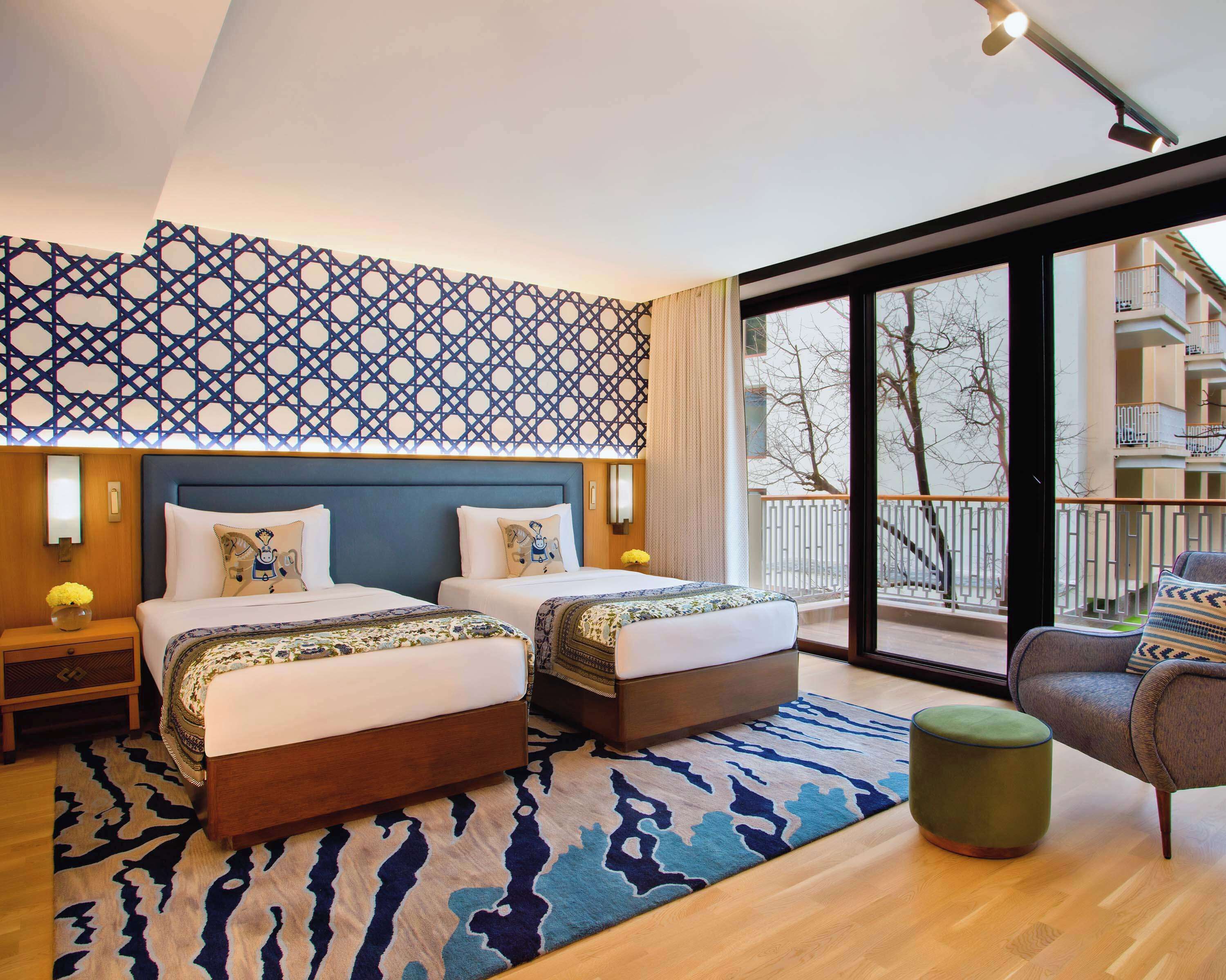 Quarto de hotel com duas camas, cabeceira estampada, uma janela grande com varanda e um banquinho verde.