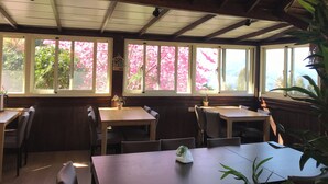 Restaurante com mesas e cadeiras de madeira, grandes janelas com flores cor-de-rosa e vista para o exterior.