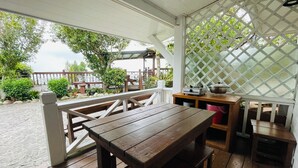 Um deck de madeira com um banco e uma mesa, cercado por um cercado de treliça branca e vegetação.
