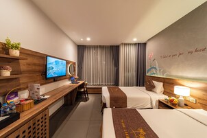 Quarto de hotel com duas camas, uma mesa com computador, uma televisão e um quadro paisagístico na parede.