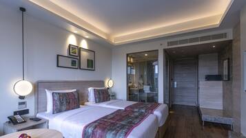 Quarto de hotel moderno com duas camas, uma mesa de cabeceira com telefone, uma mesa redonda com frutas e um banheiro visível através de uma porta aberta.