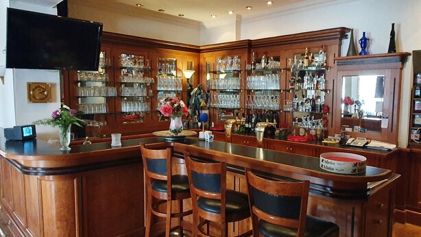 Uma área de bar com banquetas de madeira, uma televisão montada acima do balcão e prateleiras repletas de copos e garrafas.