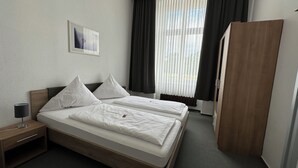 Quarto de hotel com duas camas, uma mesinha de cabeceira, uma luminária, uma janela com cortinas e um guarda-roupa de madeira.