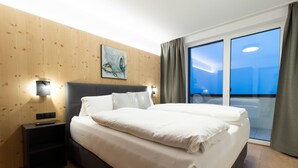 Quarto de hotel moderno com uma cama grande, painéis de madeira, uma pintura e vista através de uma porta de vidro.