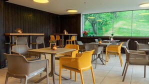 Restaurante com várias mesas e cadeiras, grandes janelas com vista para o verde e uma parede de madeira.