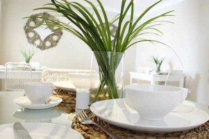 Mesa de jantar posta com pratos, tigelas e talheres brancos, acompanhada por um vaso de planta.