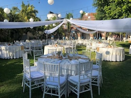 Uma recepção de casamento ao ar livre com mesas redondas, cadeiras brancas e luzes de fada.