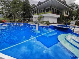 Uma piscina com água azul cristalina, rodeada por uma área ajardinada com árvores e um edifício ao fundo.