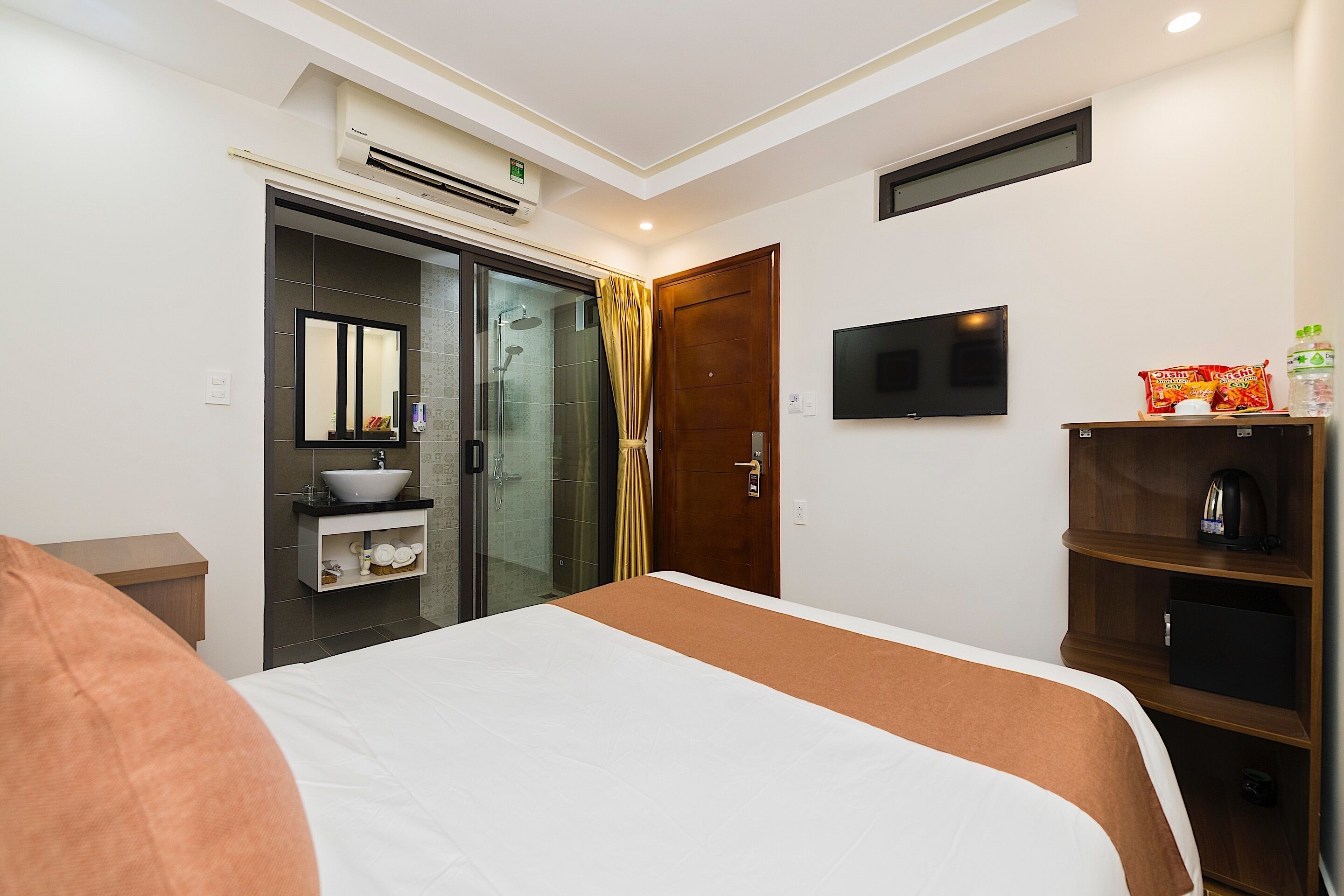 Quarto de hotel com cama, banheiro com chuveiro de vidro, televisão e prateleira com artigos de higiene pessoal.