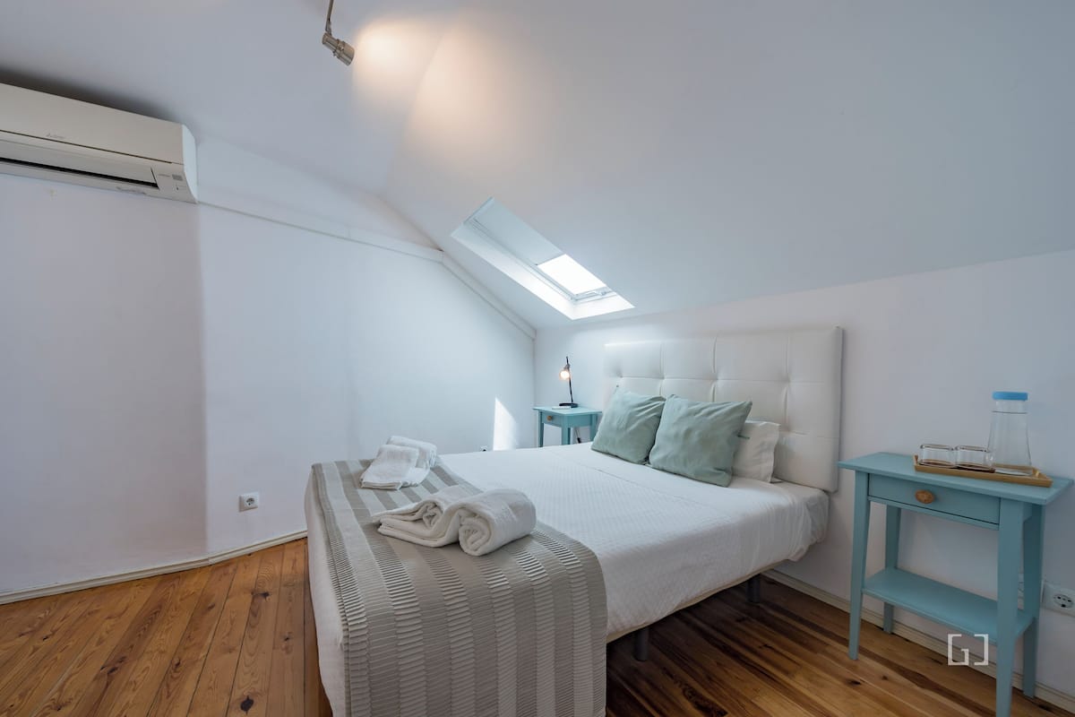 Quarto com claraboia, cama com roupa de cama branca, mesa de cabeceira com abajur e uma pequena mesinha de noite.