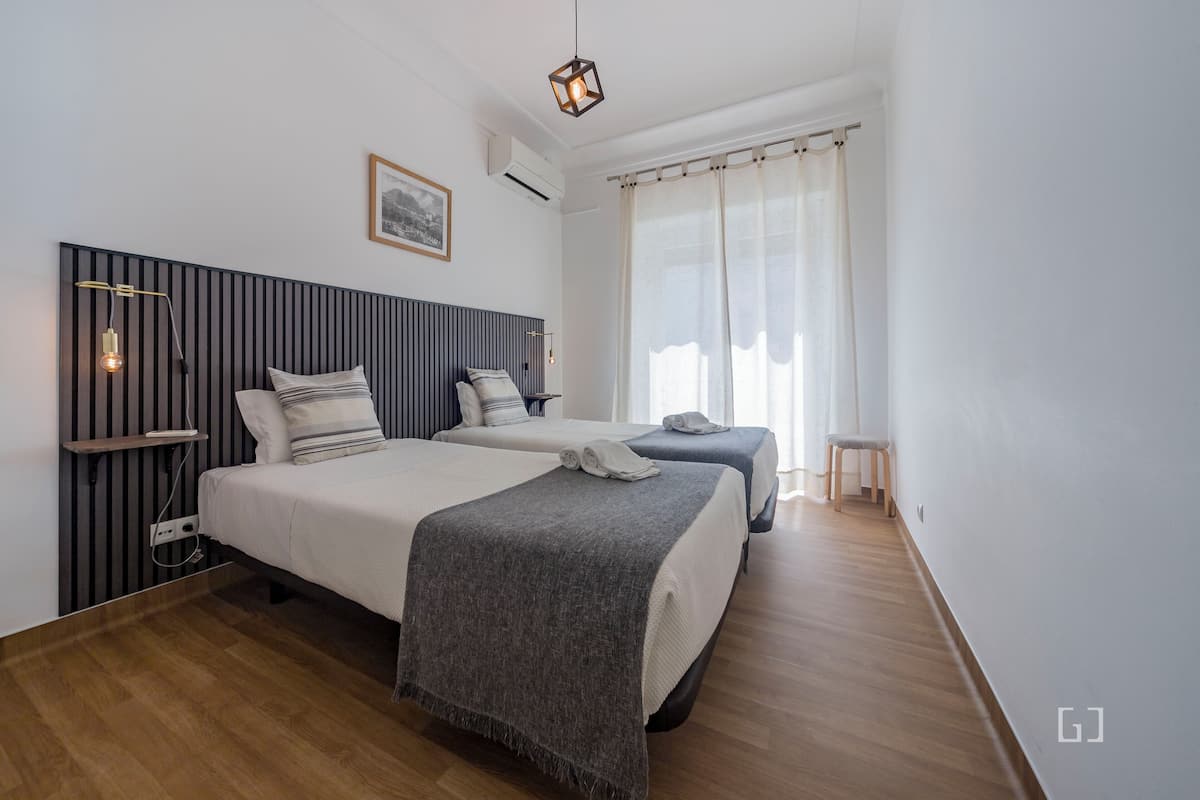 Quarto de hotel moderno com duas camas, uma mesinha de cabeceira, um banquinho pequeno e um lustre.