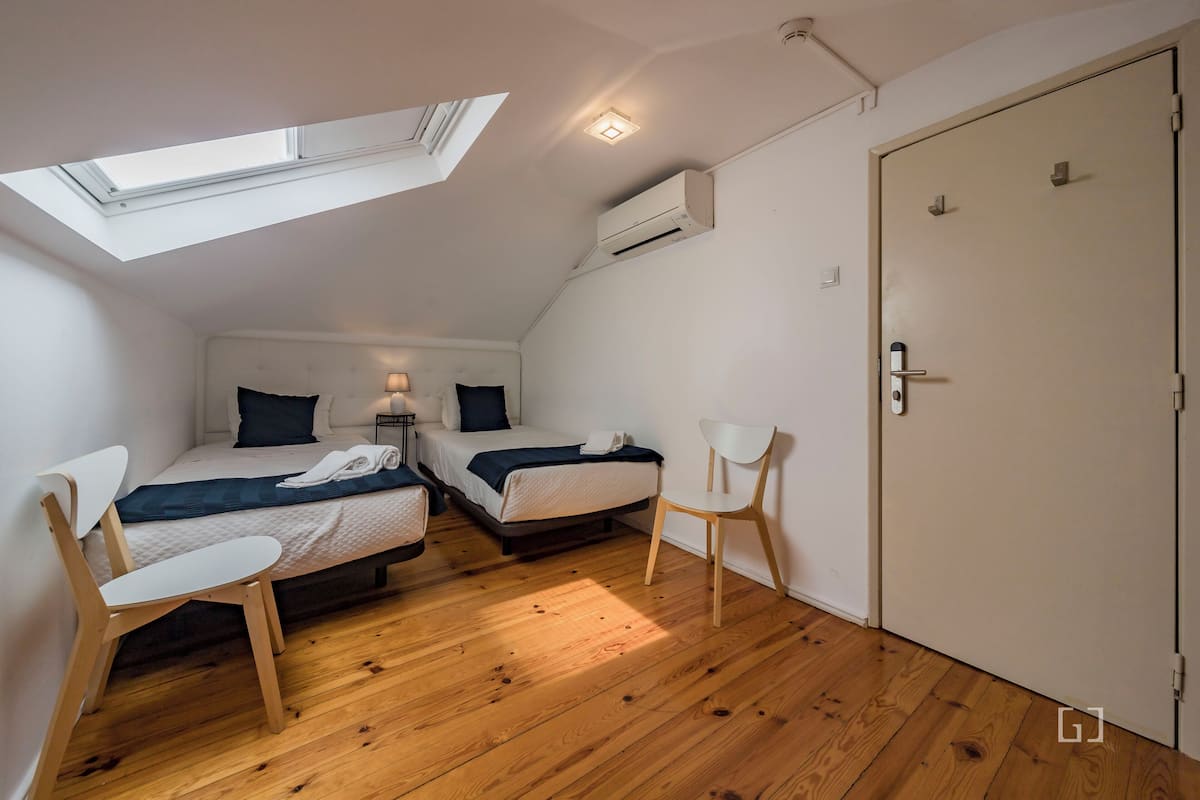 Quarto pequeno e bem iluminado com duas camas, um claraboia, uma porta e piso de madeira.