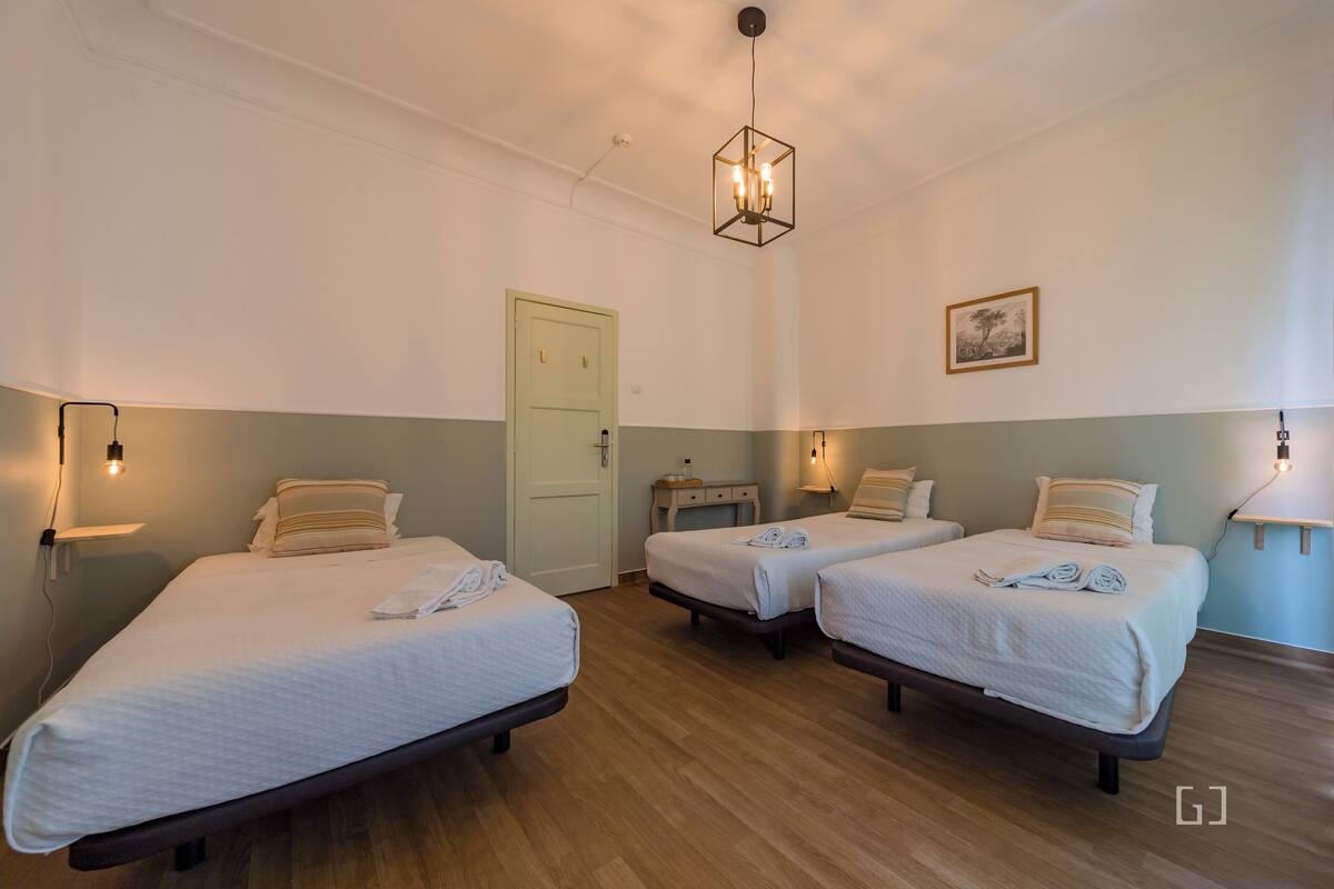 Quarto de hotel com três camas de solteiro, piso de madeira, porta verde e um quadro na parede.