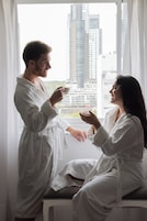 Um casal de roupões brancos senta-se junto a uma janela com vista para a cidade.