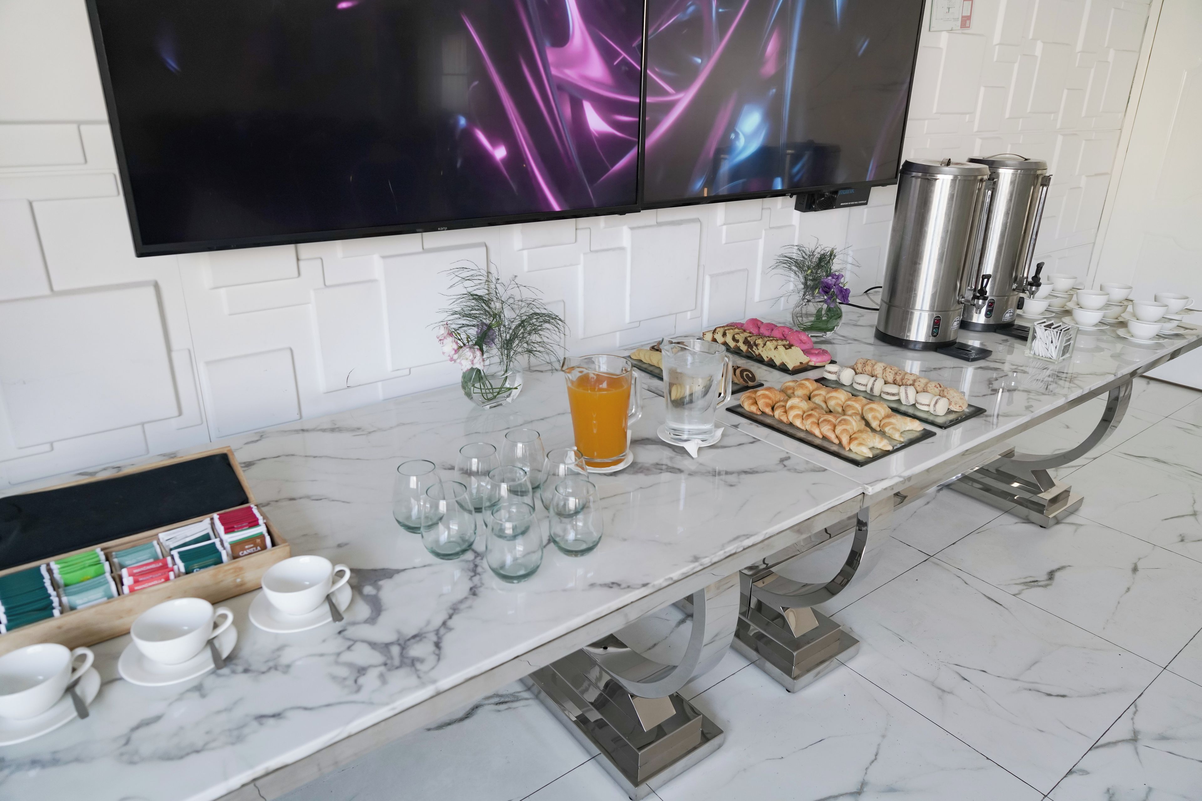 Uma área de bar com bancada de mármore, TV de tela plana, máquinas de café e diversos pastéis e bebidas.