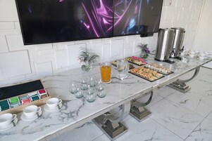 Uma área de bar com bancada de mármore, TV de tela plana, máquinas de café e diversos pastéis e bebidas.