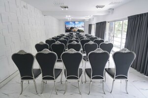 Uma sala de conferências com fileiras de cadeiras voltadas para uma tela grande, paredes brancas e uma janela com cortinas.