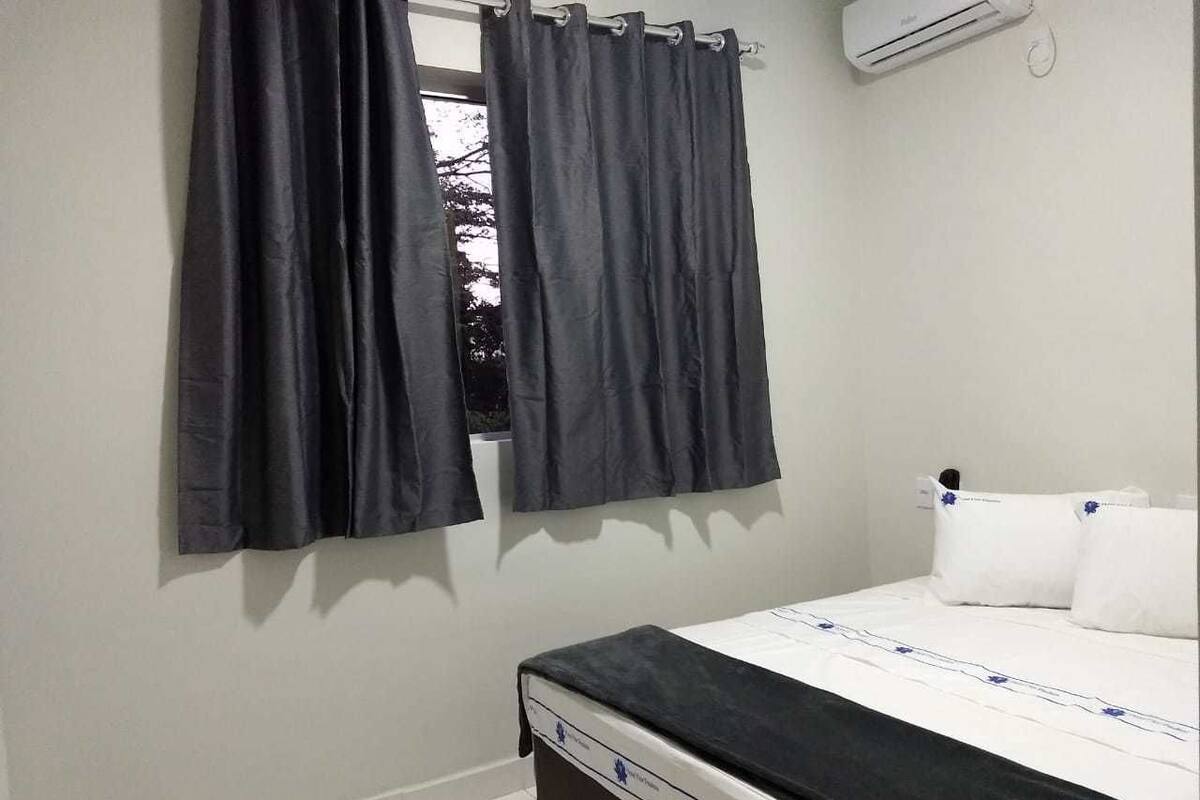 Quarto com cama, almofadas brancas, janela com cortinas escuras e um aparelho de ar condicionado na parede.