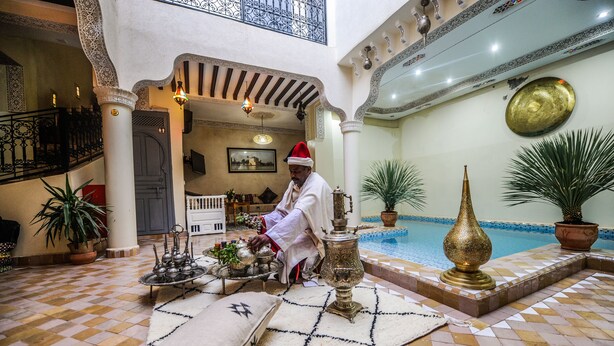 Interior luxuoso com chão em mosaico, colunas ornamentadas, área de piscina e um homem com vestes tradicionais.