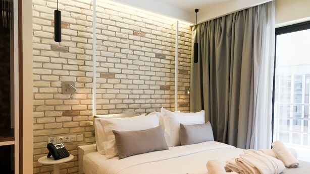 Quarto de hotel com parede de tijolos, cama com roupa de cama branca, mesa de cabeceira com telefone e janela com cortinas.