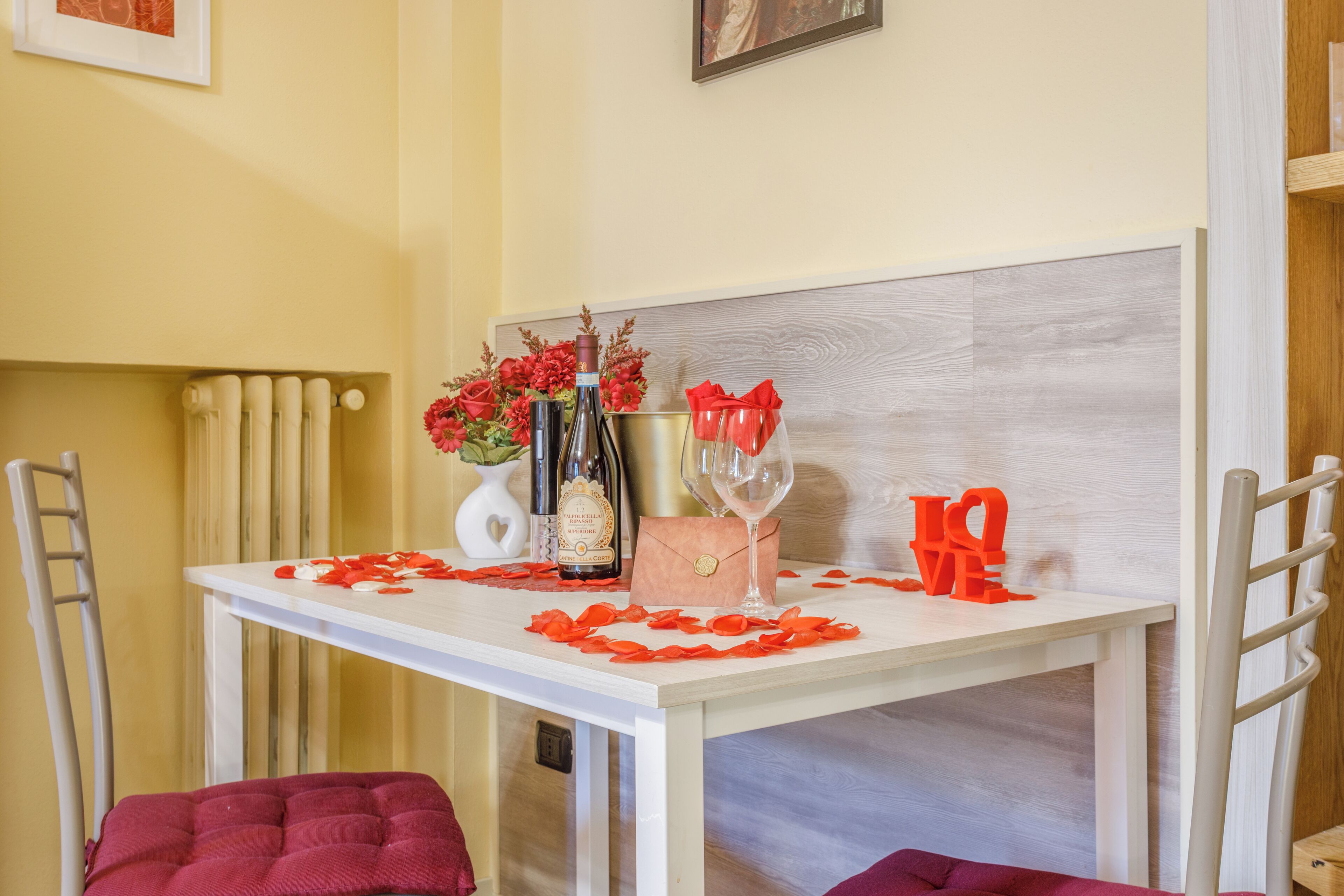 Uma mesa de jantar com toalha branca, pétalas de rosa vermelhas, uma garrafa, um copo e uma decoração em formato de coração vermelho.