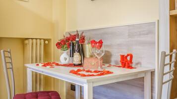 Uma mesa de jantar com toalha branca, pétalas de rosa vermelhas, uma garrafa, um copo e uma decoração em formato de coração vermelho.