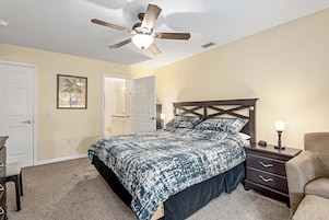 Quarto com cama, mesa de cabeceira, cadeira, ventoinha no teto e porta.