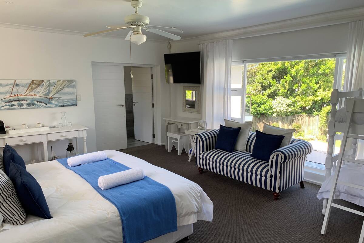 Quarto de hotel com cama, sofá riscado, escrivaninha com cadeira, televisão e janela com vista para vegetação.