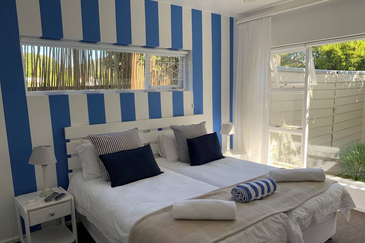 Uma cama de casal com roupa de cama branca, duas almofadas, uma mesa de cabeceira com abajur e uma janela com vista para o verde.