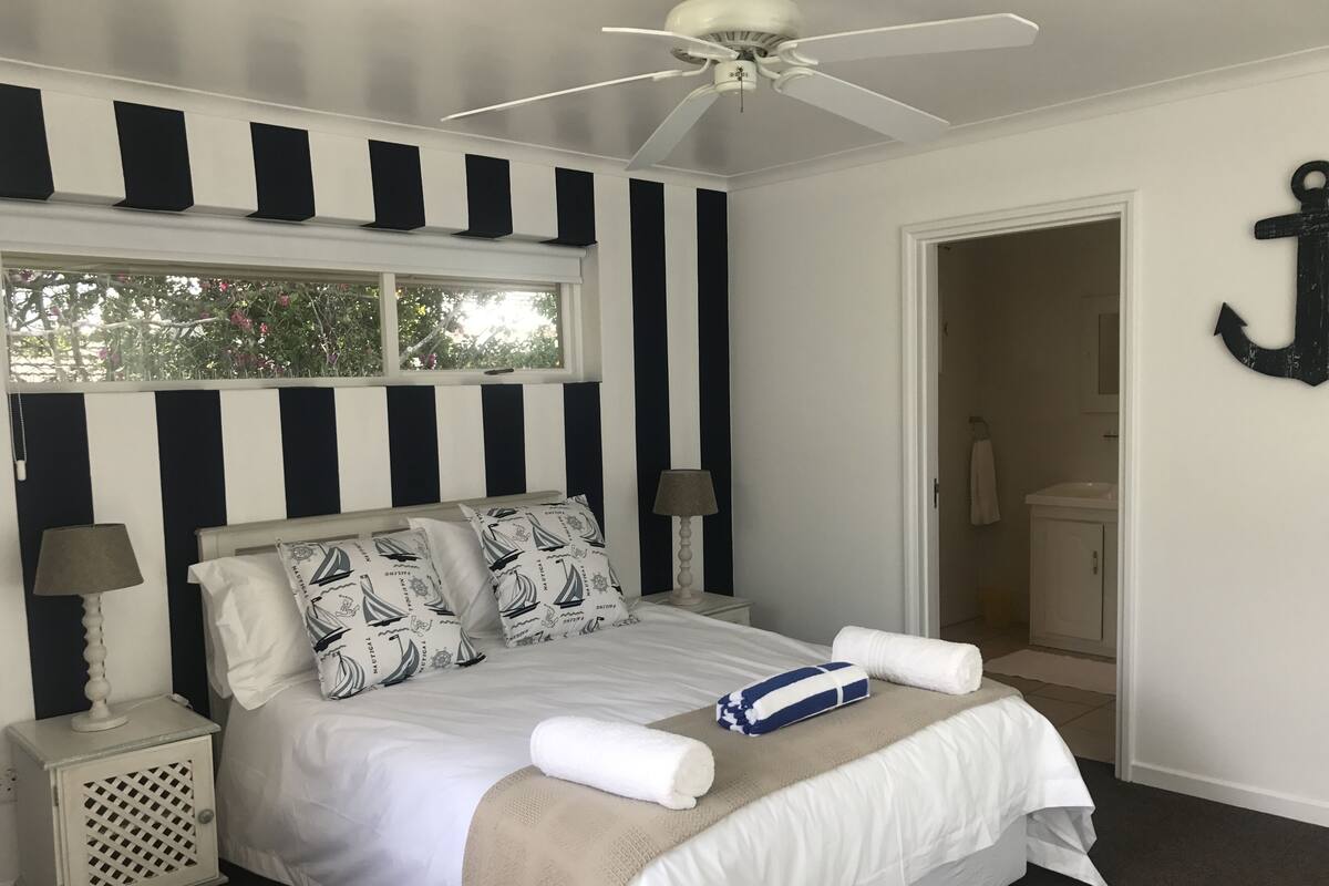 Cama bem arrumada com cabeceira listrada, abajur de cabeceira e janela com vista para o verde.