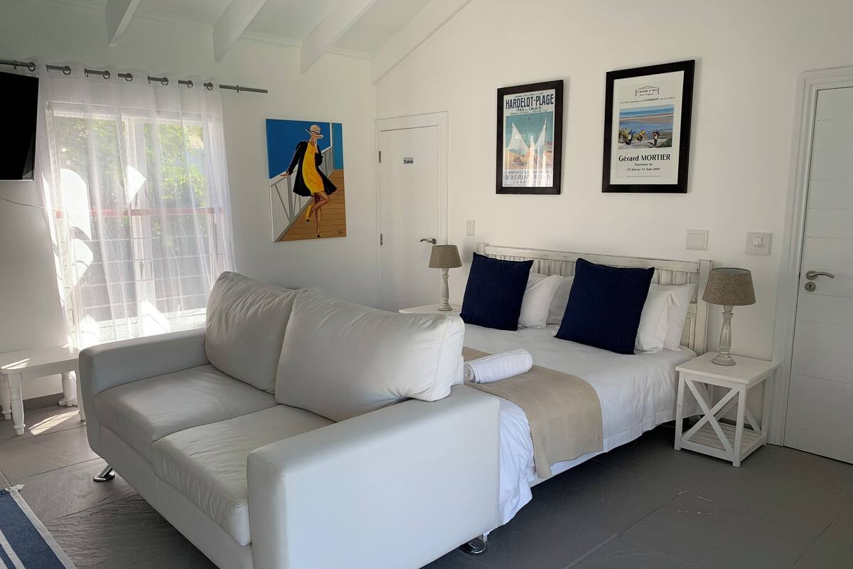 Quarto de hotel moderno com um sofá branco, uma cama com almofadas azul-escuras, uma pequena mesa de cabeceira com uma luminária e dois cartazes emoldurados na parede.