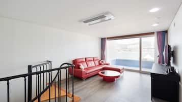 Uma sala de estar moderna com um sofá vermelho, uma mesa de centro redonda e uma escrivaninha preta. Há uma escada com guarda-corrimão preto e corrimão de madeira. O quarto possui grandes janelas com cortinas e vista para a cidade.