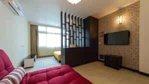 Quarto de hotel moderno com um sofá vermelho grande, uma televisão de tela plana na parede, uma pequena escrivaninha e um painel decorativo.