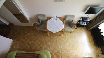 Quarto com assoalho de madeira parquete, uma poltrona verde, uma mesa redonda pequena com toalha branca, duas poltronas, uma televisão de tela plana em um suporte e um quadro na parede.