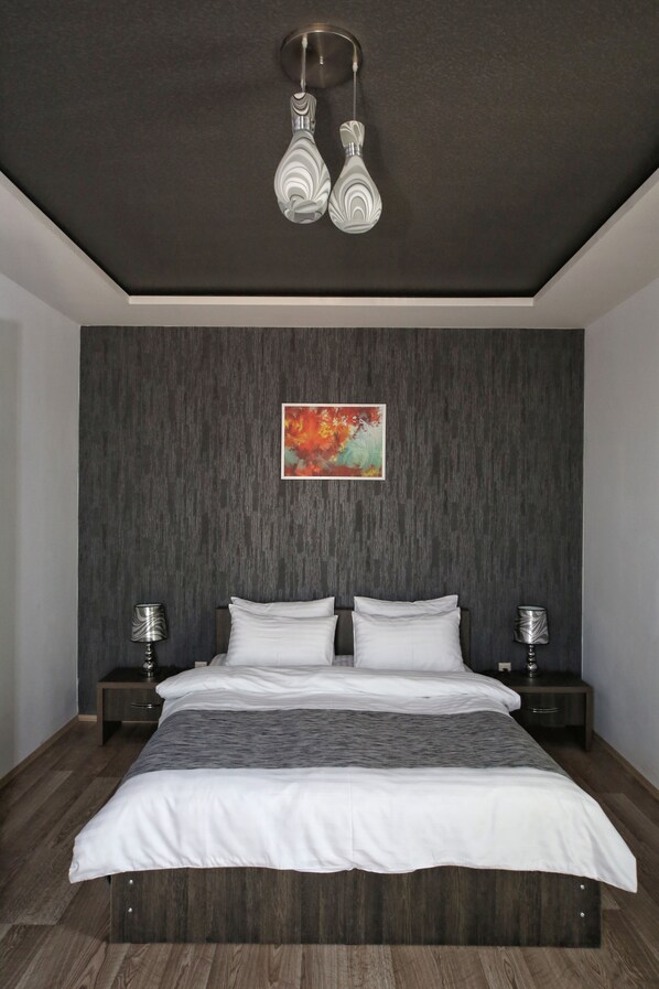 Quarto moderno com uma cama grande, piso de madeira escura e uma parede cinza texturizada. Há uma pintura sobre a cama e duas luminárias de cabeceira.