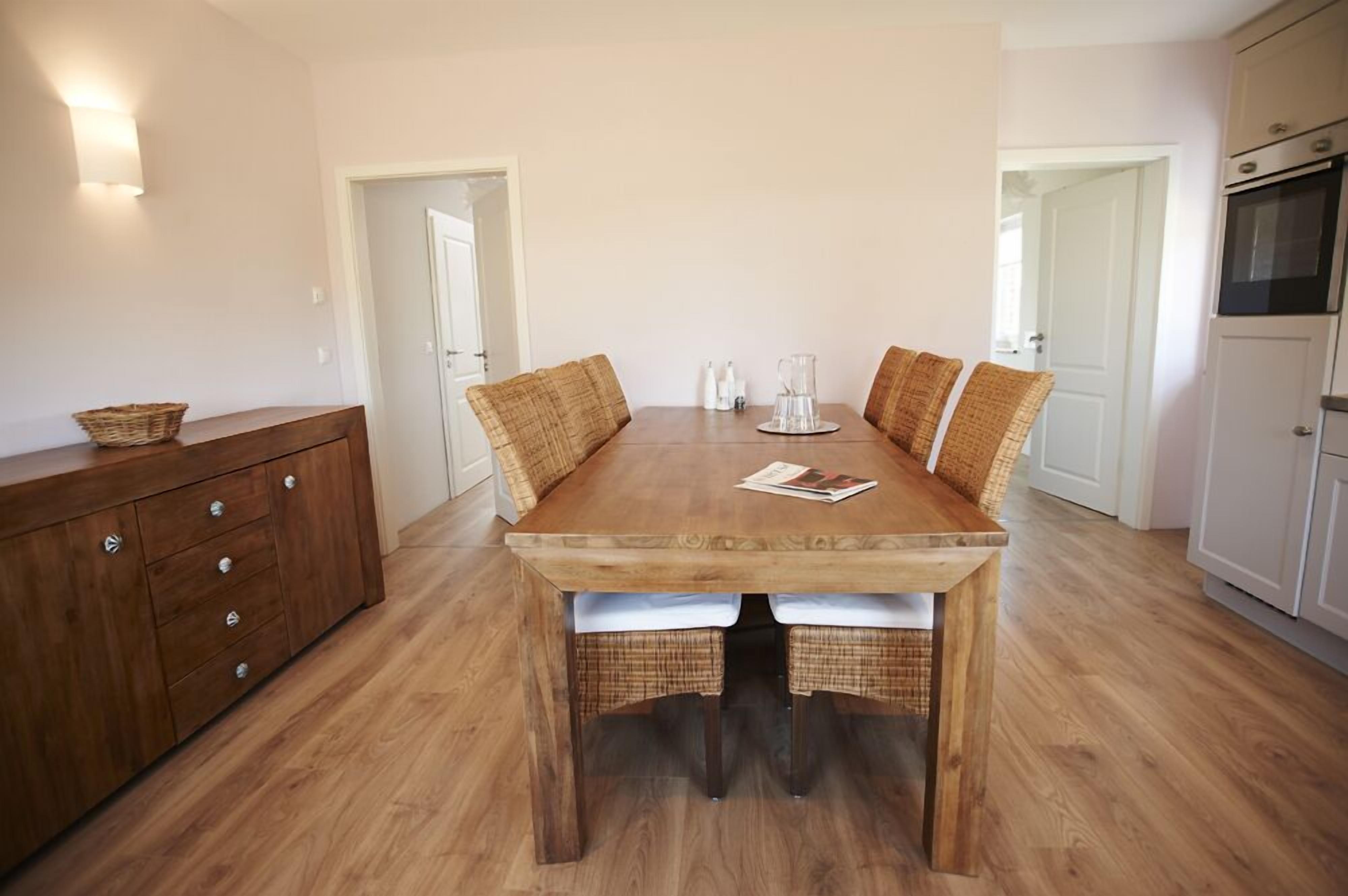 Uma sala de jantar com uma mesa de madeira, quatro cadeiras, um armário de madeira e uma cozinha ao fundo.