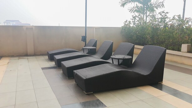 Cadeiras de descanso ao ar livre e uma pequena mesa num pátio ladrilhado, com vista para um edifício e vegetação ao fundo.